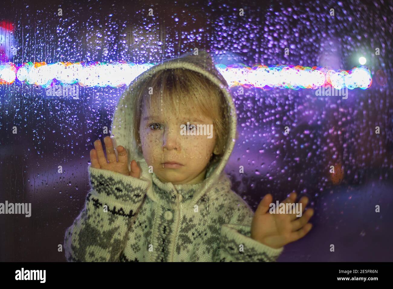 Little child, toddler boy, standing behind window with drops outdoors ...