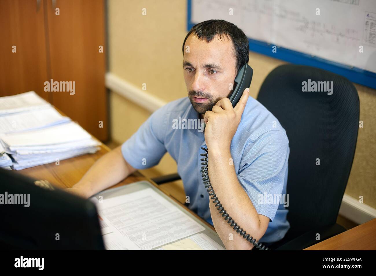A young employee sits in a chair at a desk in the office and talks on a ...