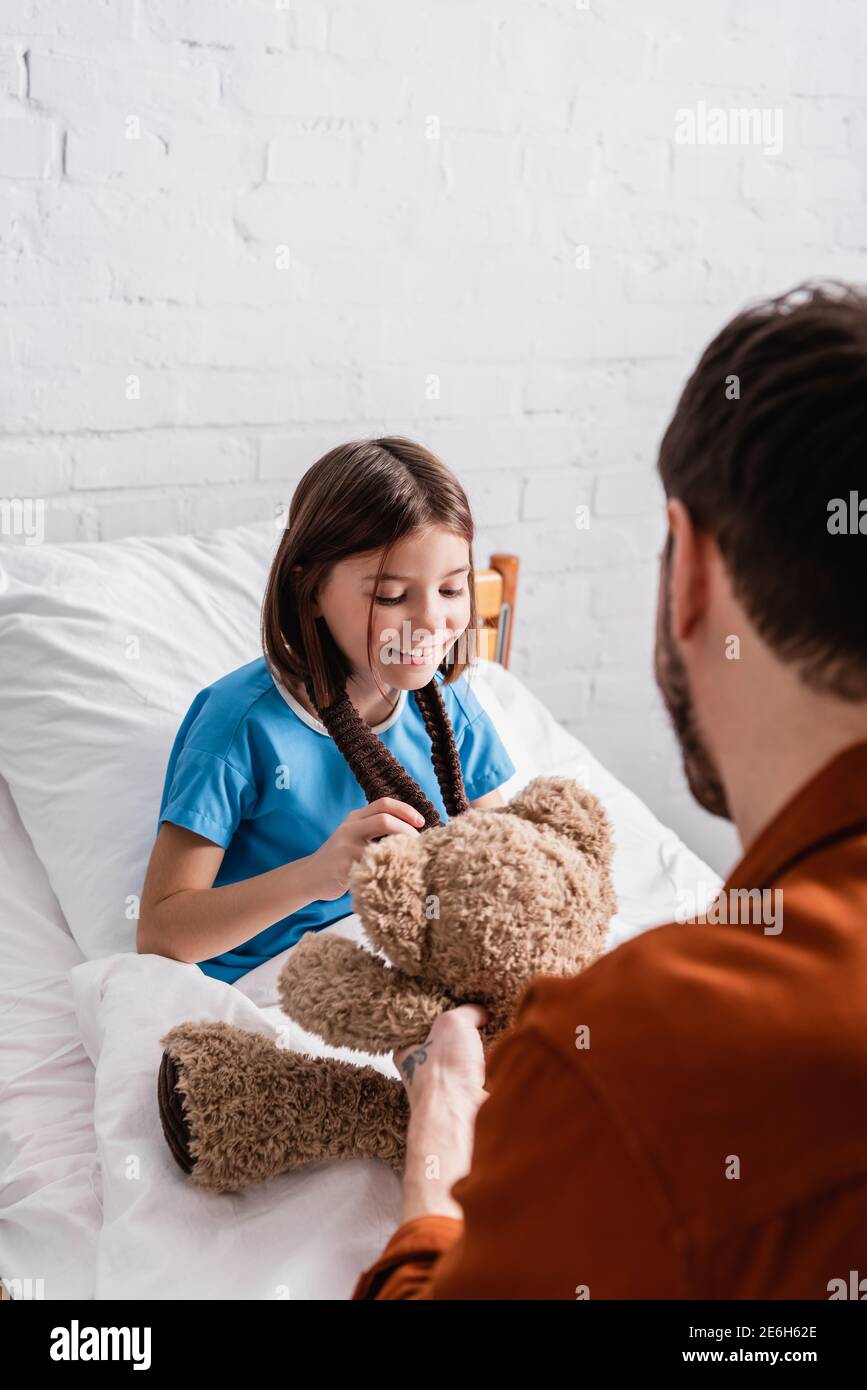happy girl touching teddy bear near father on blurred foreground in ...