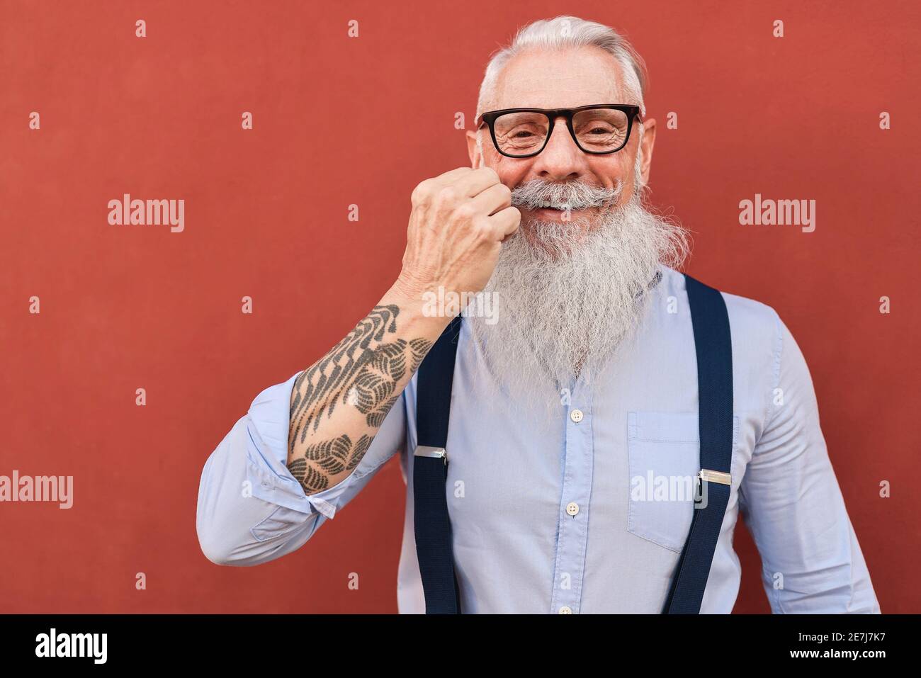 Happy hipster senior man touching mustache with red wall in background ...
