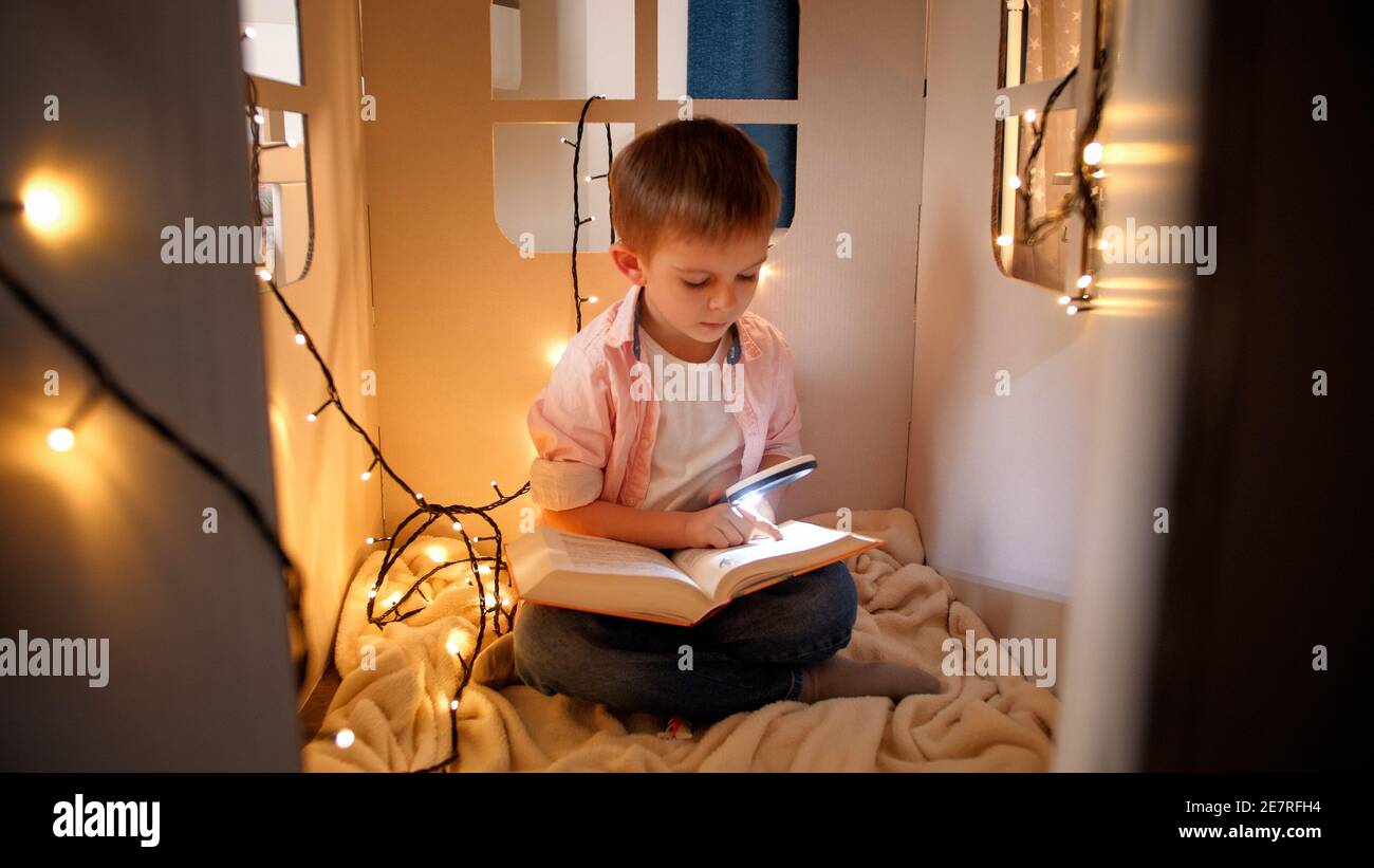 Cute little boy reading book with torch in his toy cardboard house at ...