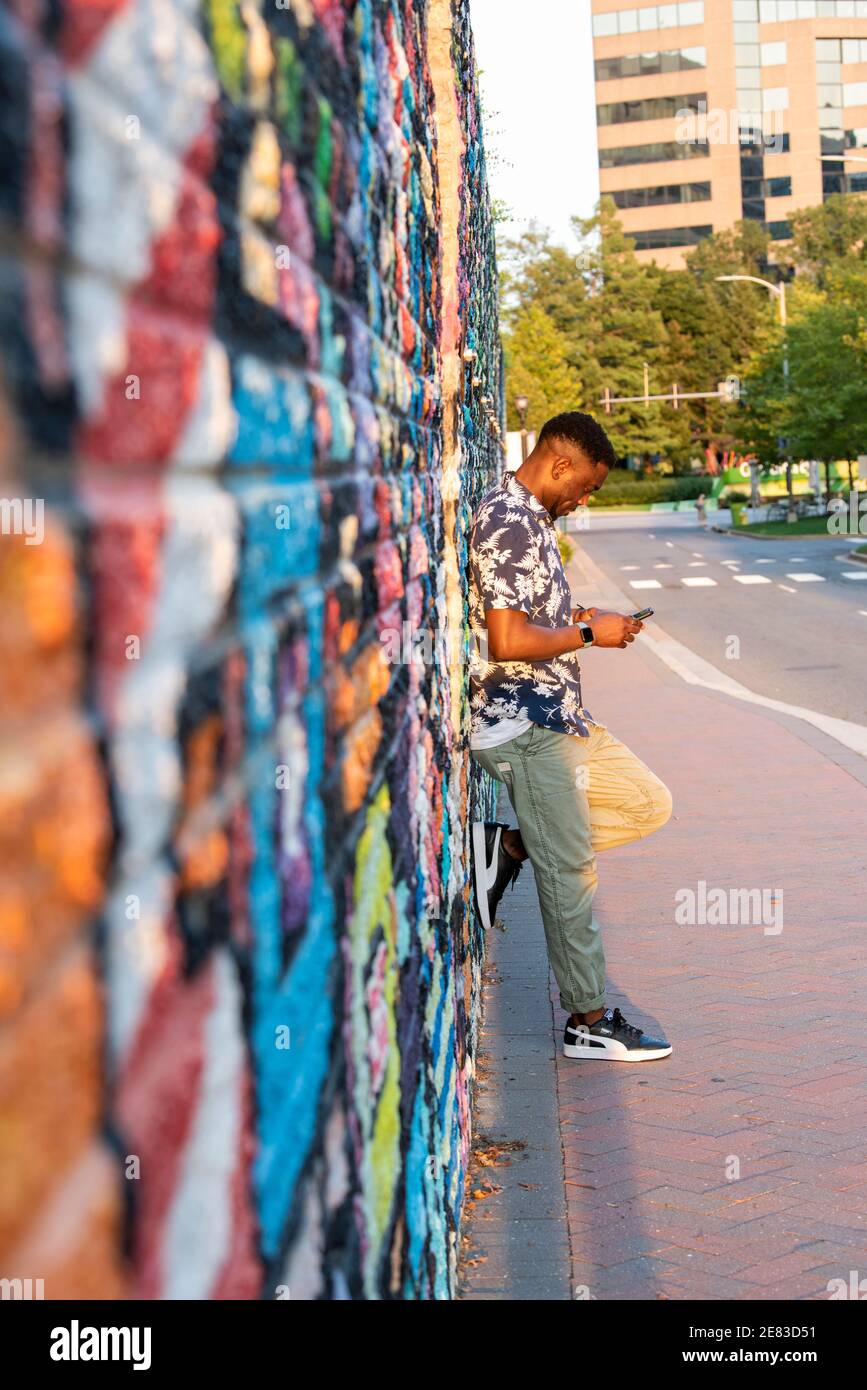 A Black man leans against a colorful wall mural and uses a cell phone ...