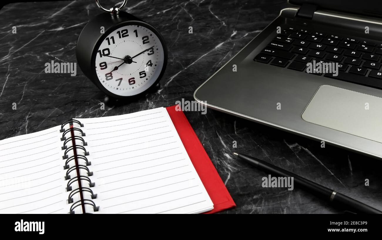 A laptop, clock and notebook on the black rough marble surface. Time ...