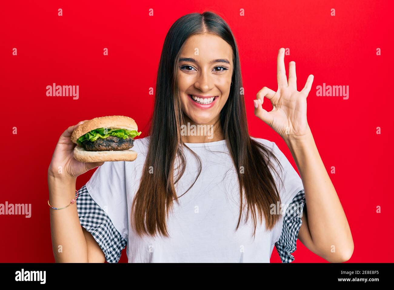 Young hispanic woman eating hamburger doing ok sign with fingers ...