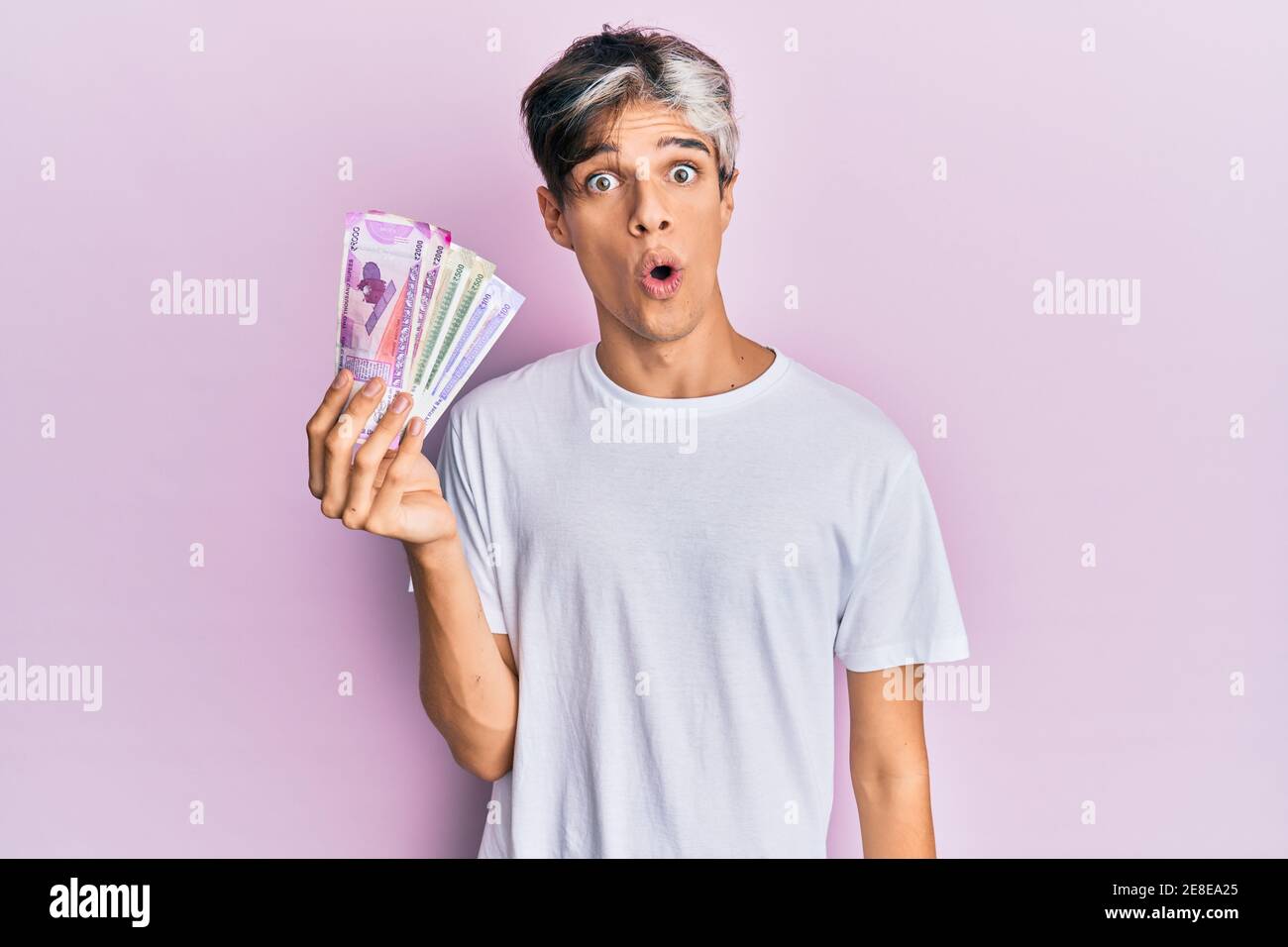 Young hispanic man holding indian rupee banknotes scared and amazed ...