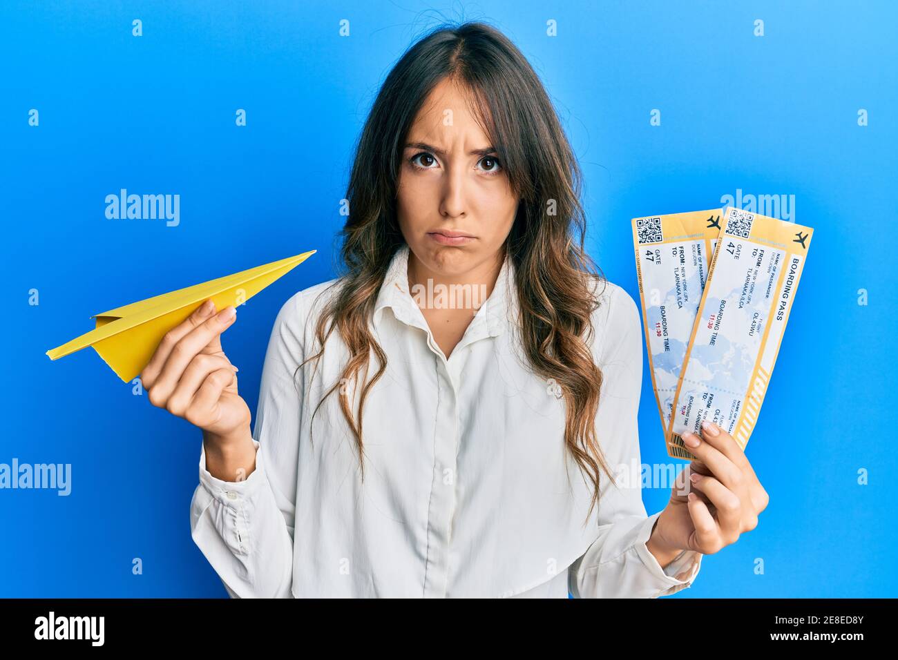 Young brunette woman holding paper airplane and boarding pass skeptic ...
