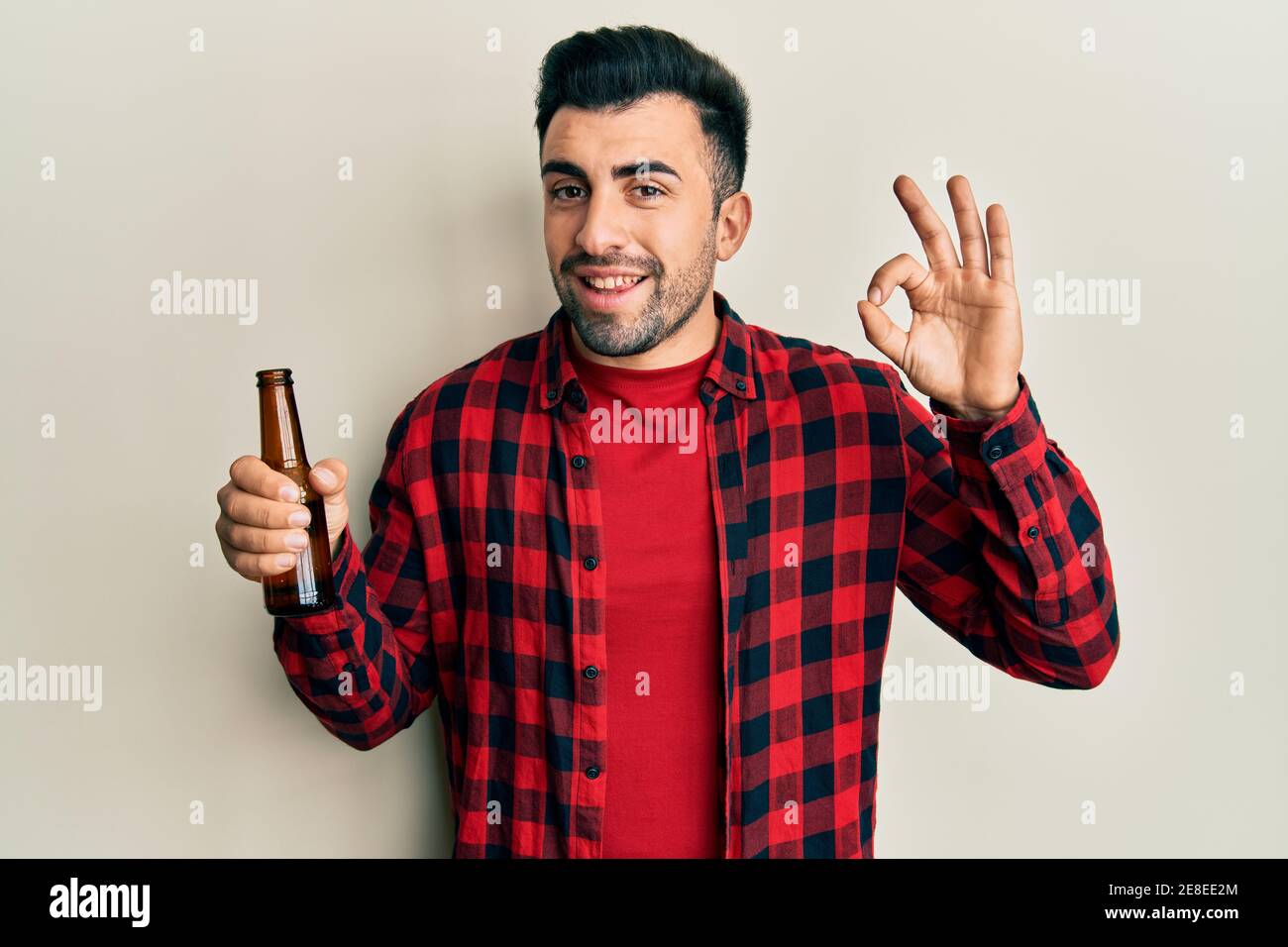 Young hispanic man drinking a bottle of beer doing ok sign with fingers ...