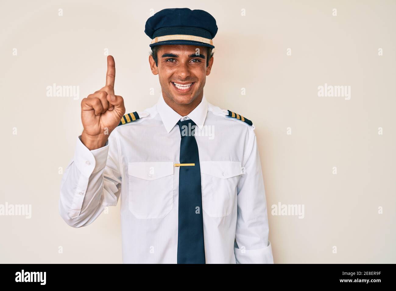 Young hispanic man wearing airplane pilot uniform pointing finger up ...