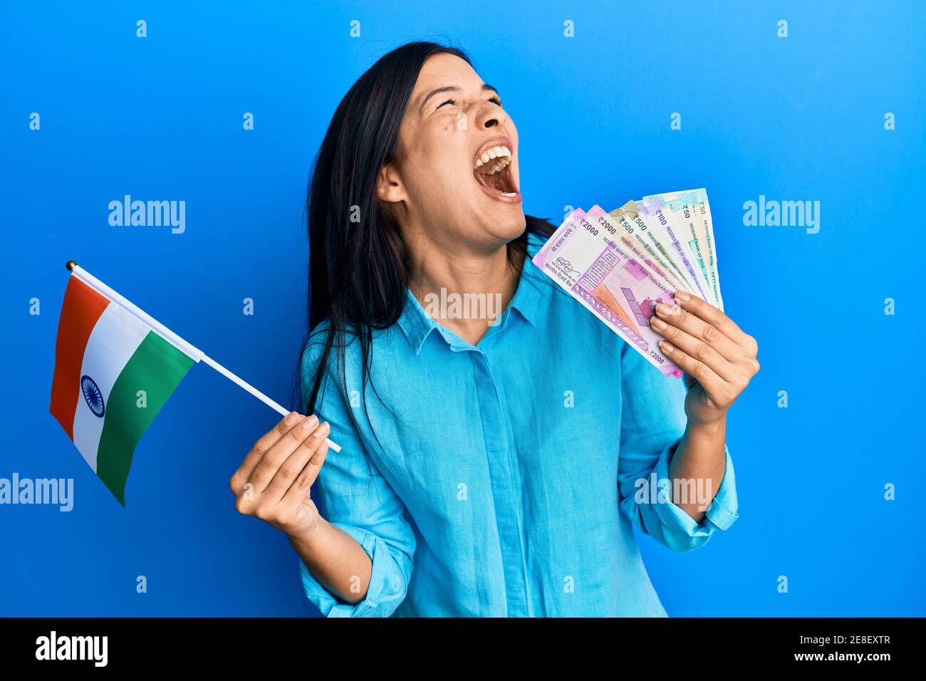 Young latin woman holding india flag and rupee banknotes angry and mad ...
