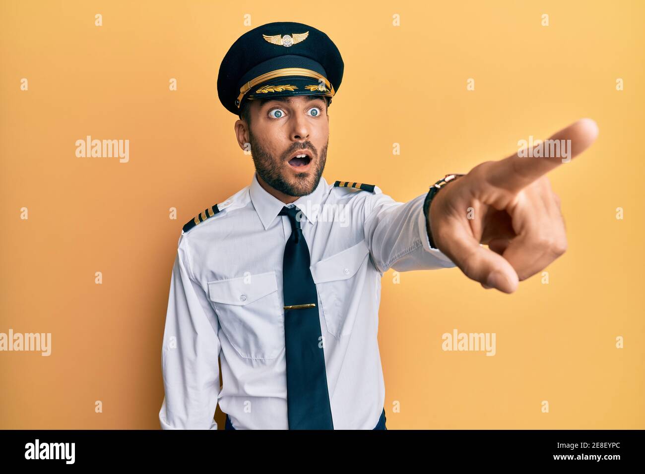 Handsome hispanic man wearing airplane pilot uniform pointing with ...