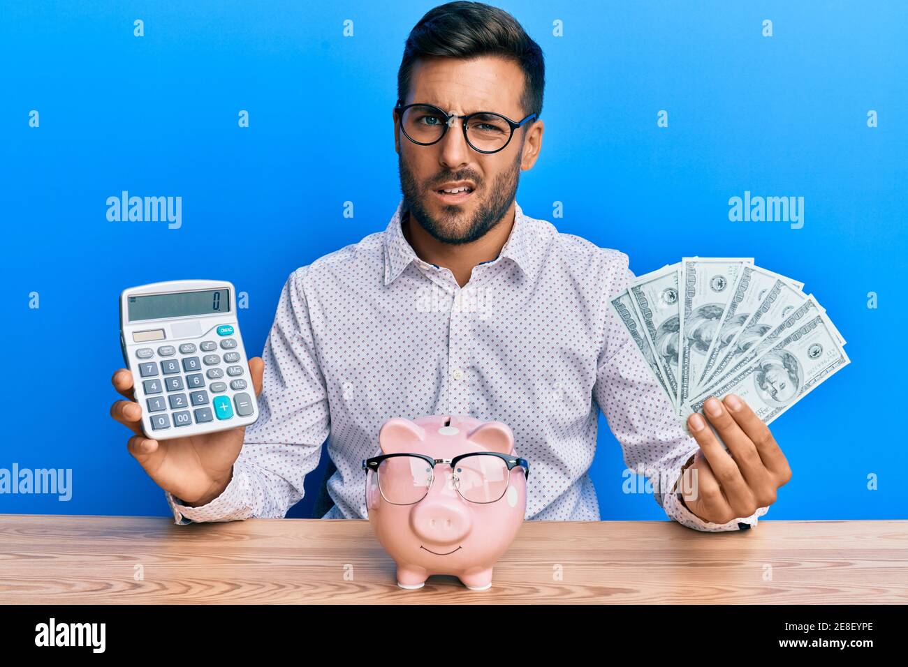Handsome hispanic man holding dollars calculating savings clueless and ...