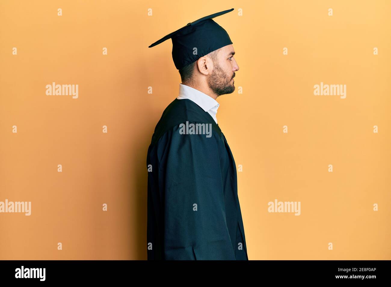 Young hispanic man wearing graduation cap and ceremony robe looking to ...
