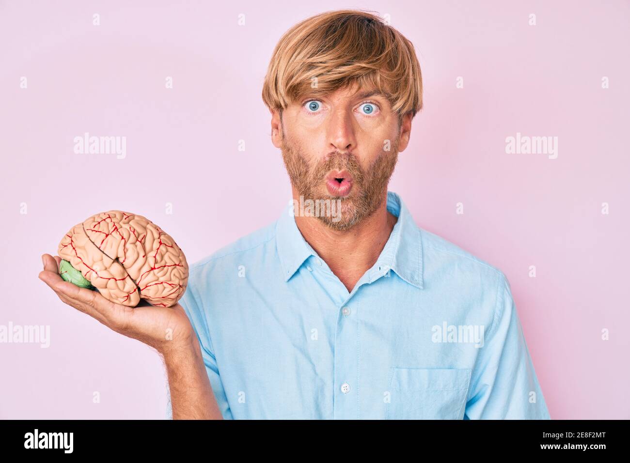 Young blond man holding brain scared and amazed with open mouth for ...