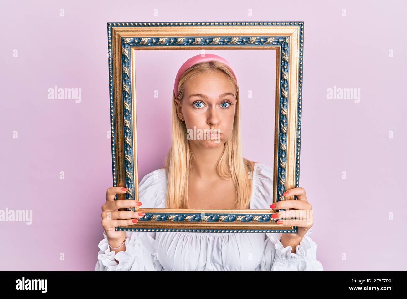Young blonde girl holding empty frame puffing cheeks with funny face ...