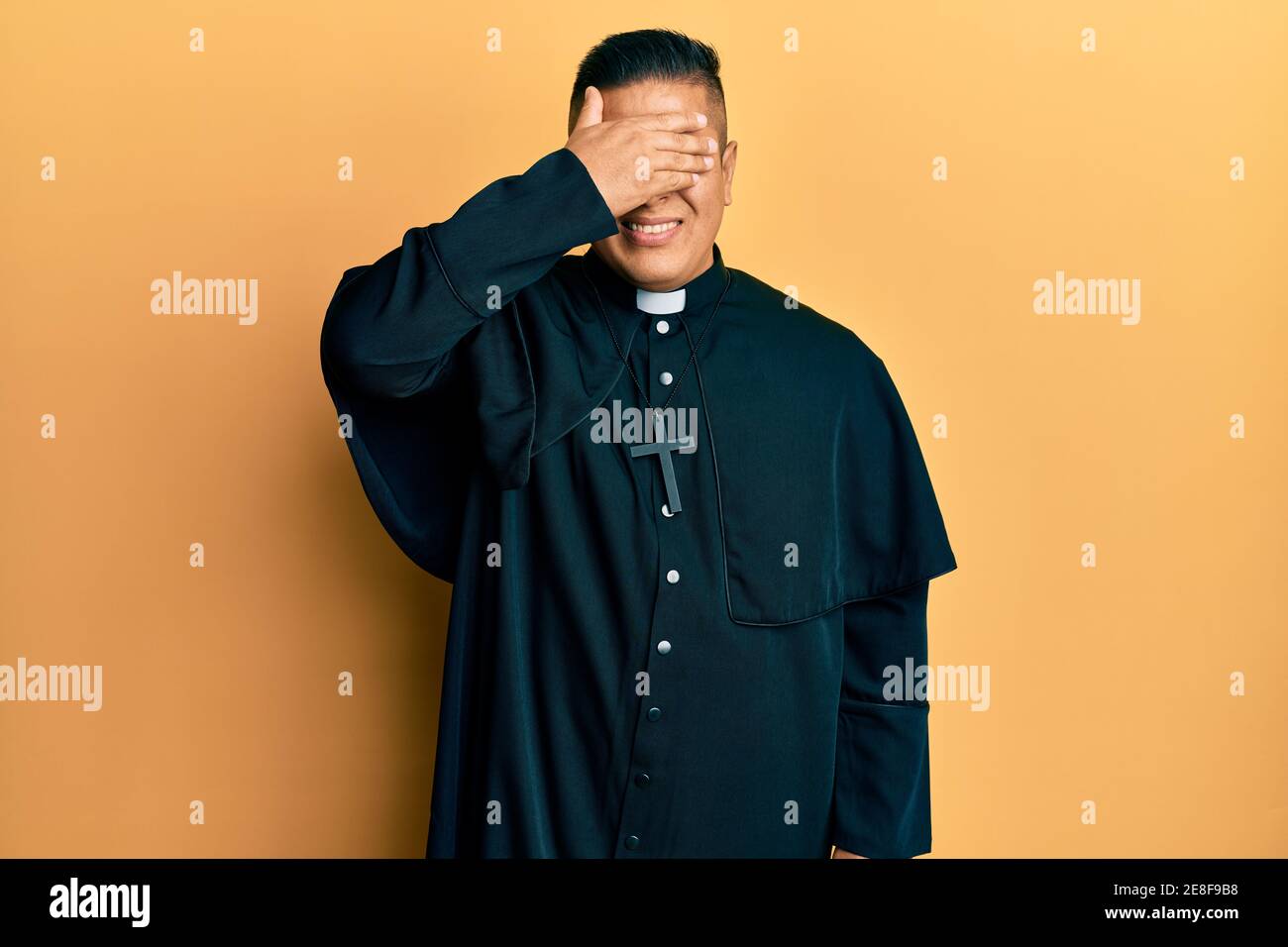 Young latin priest man standing over yellow background smiling and ...
