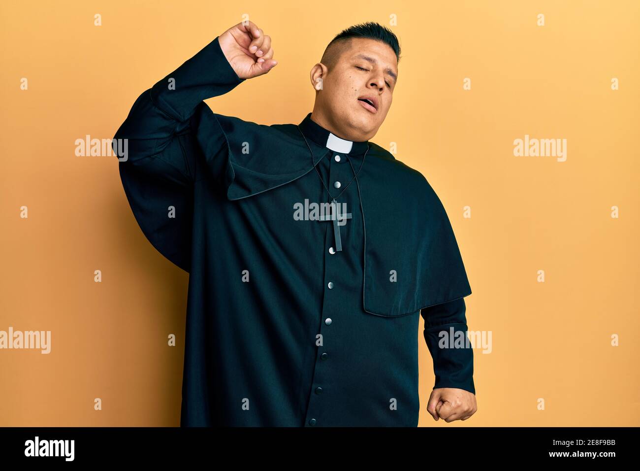 Young latin priest man standing over yellow background stretching back ...