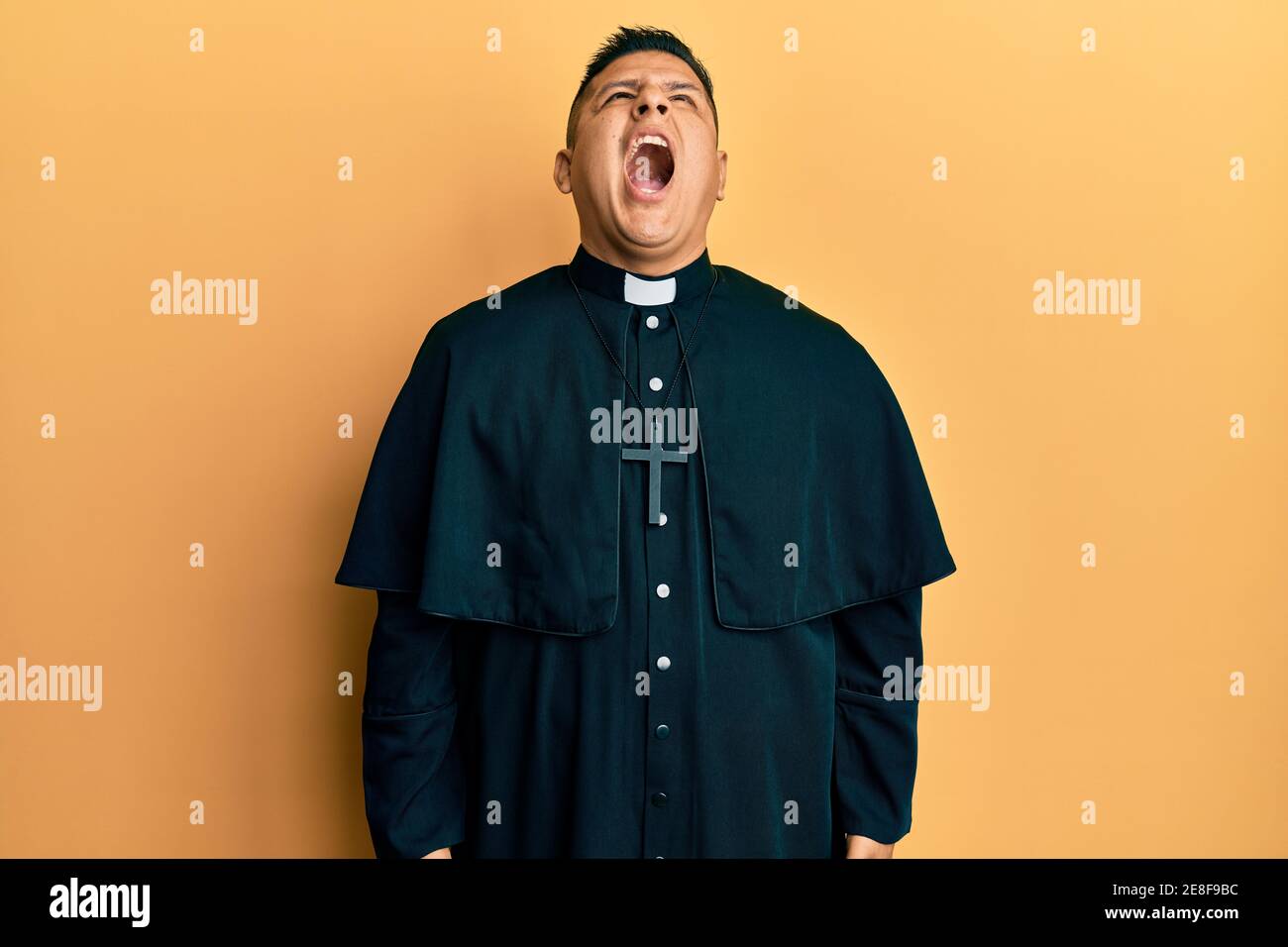 Young latin priest man standing over yellow background angry and mad ...