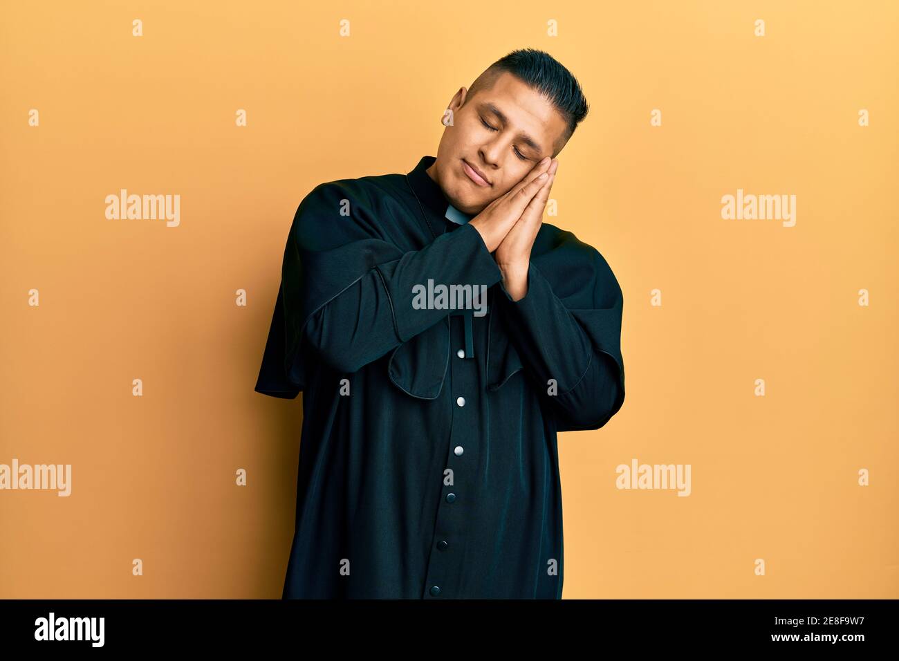 Young latin priest man standing over yellow background sleeping tired ...