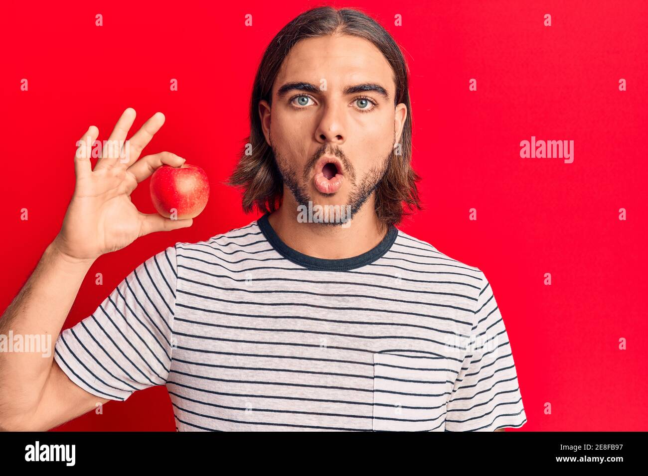 Young handsome man holding red apple scared and amazed with open mouth ...
