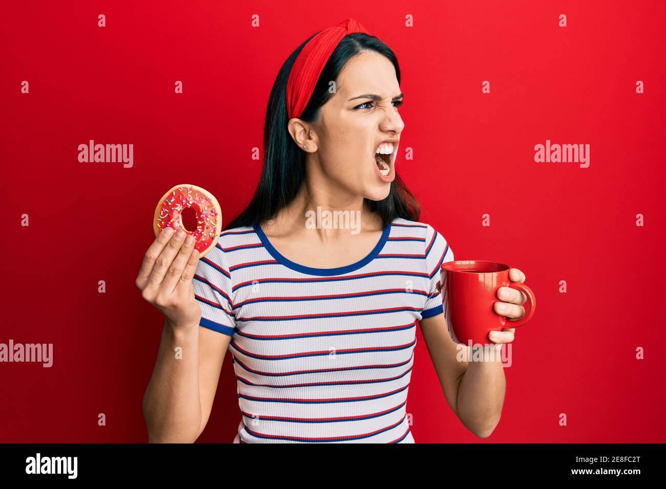 Young hispanic woman eating doughnut and drinking coffee angry and mad ...