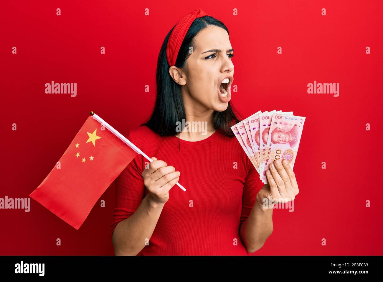 Young hispanic woman holding china flag and yuan banknotes angry and ...