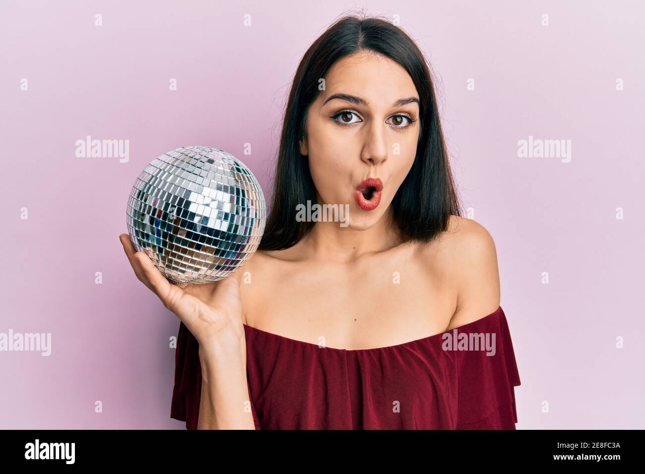 Young hispanic woman holding shiny disco ball scared and amazed with ...