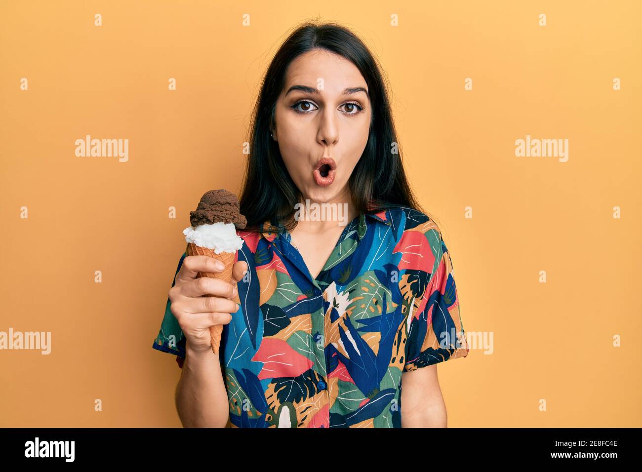 Young hispanic woman holding ice cream scared and amazed with open ...