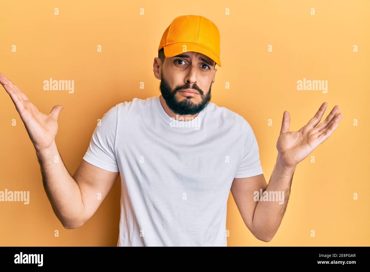 Young man with beard wearing yellow cap clueless and confused with open ...