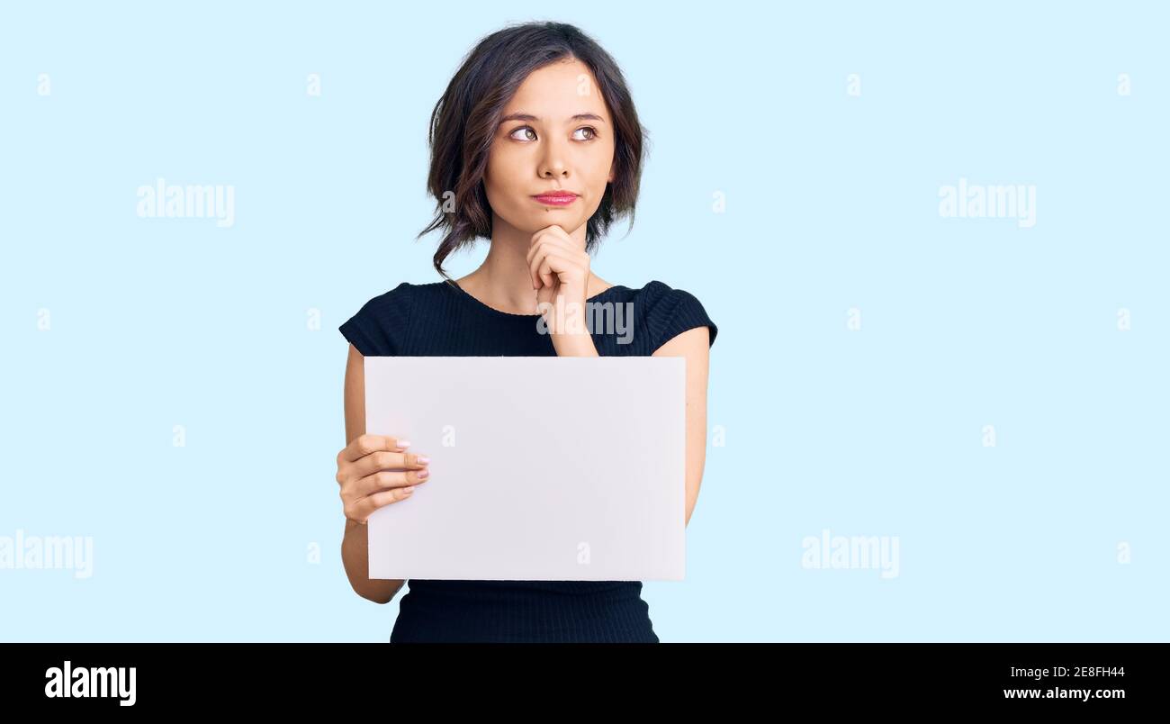 Young beautiful girl holding blank empty banner serious face thinking ...