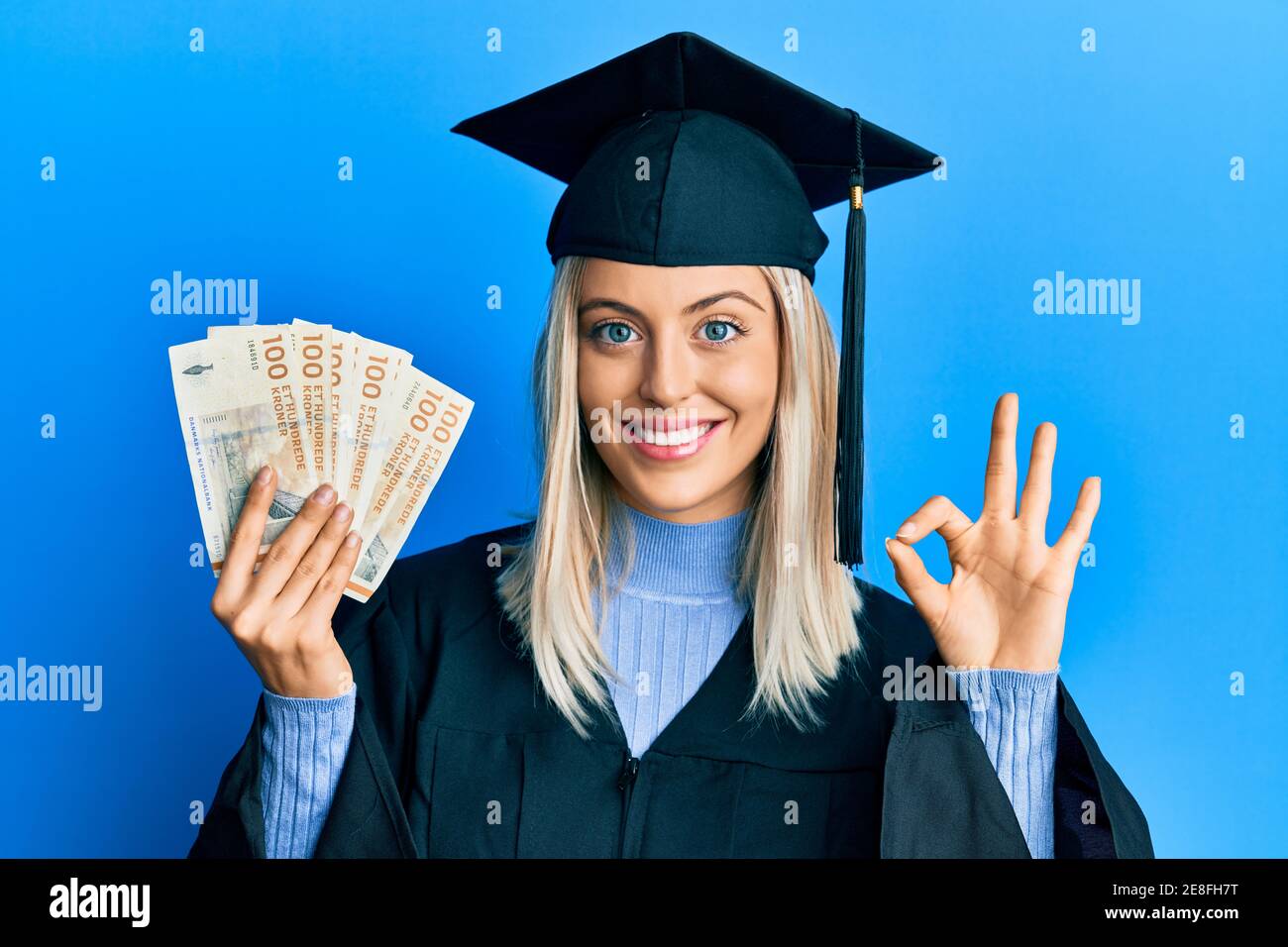 Beautiful blonde woman wearing graduation cap and ceremony robe holding ...