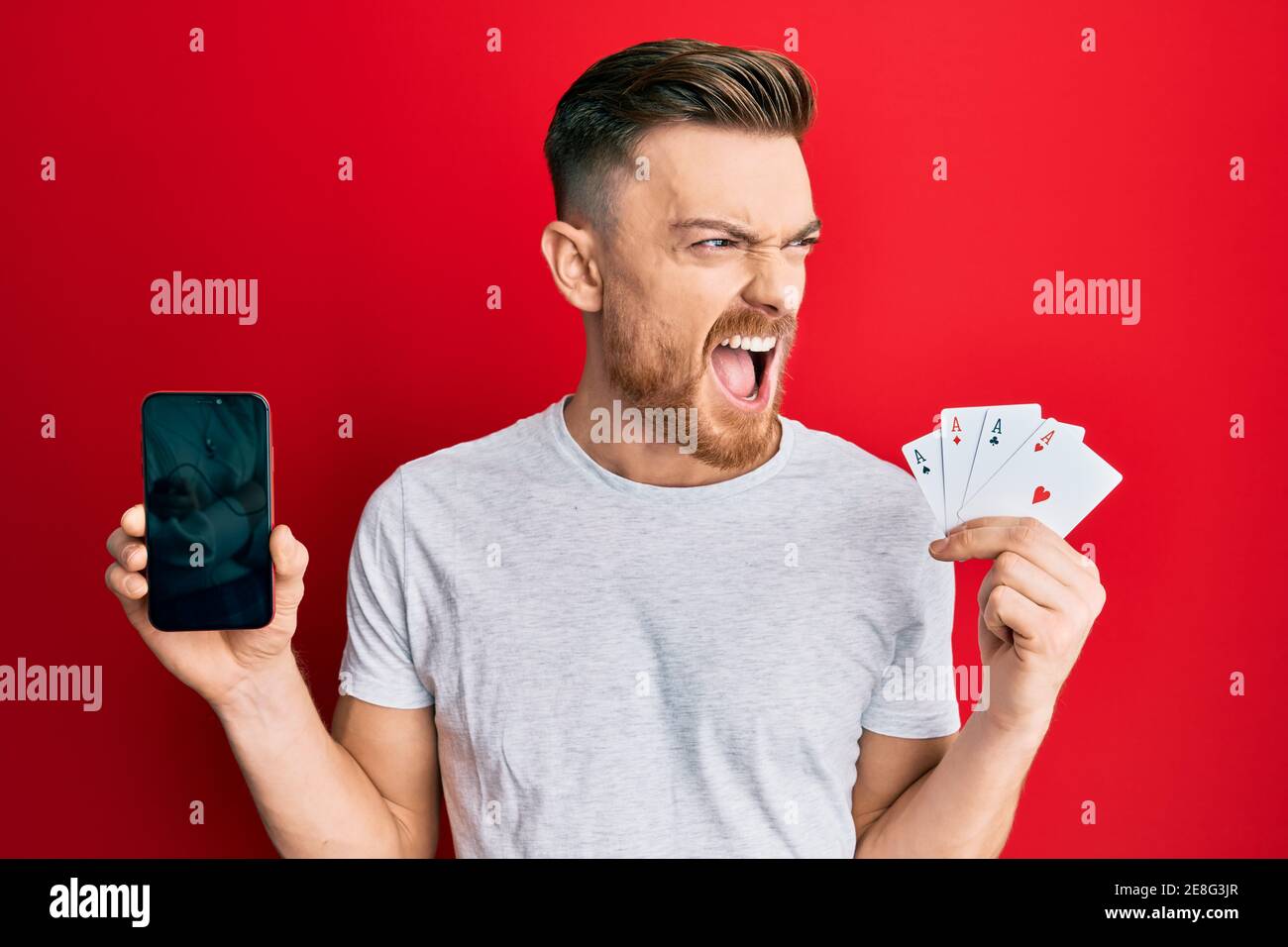 Young redhead man holding smartphone and casino card angry and mad ...