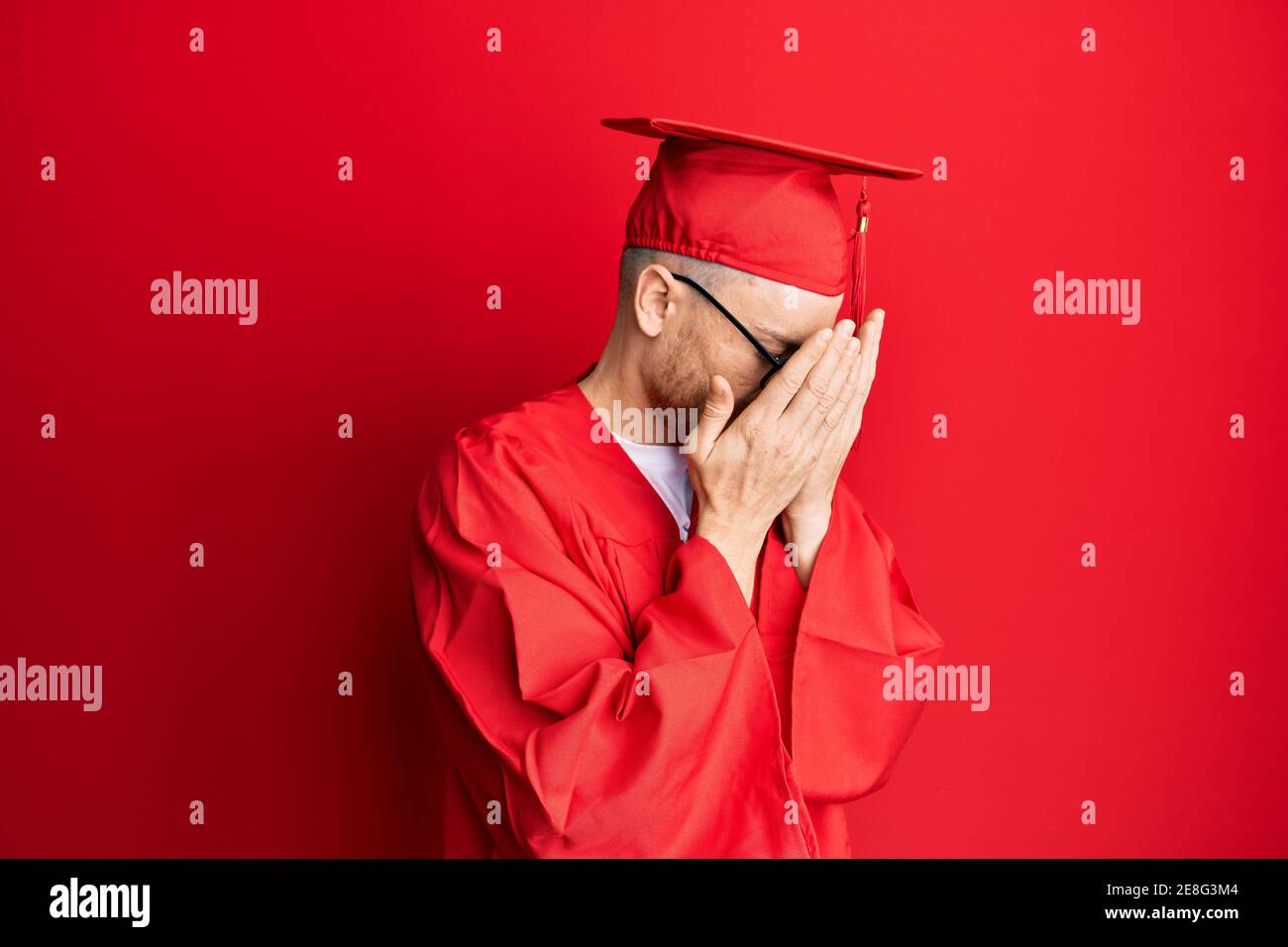 Young redhead man wearing red graduation cap and ceremony robe with sad ...