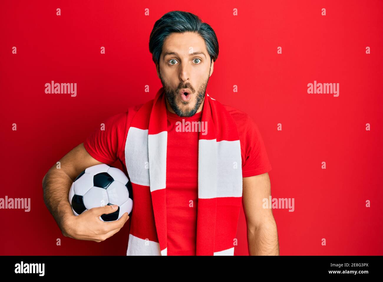 Young hispanic man football hooligan holding ball scared and amazed ...