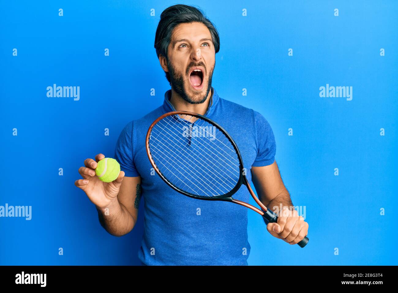 Young hispanic man playing tennis holding racket and ball angry and mad ...