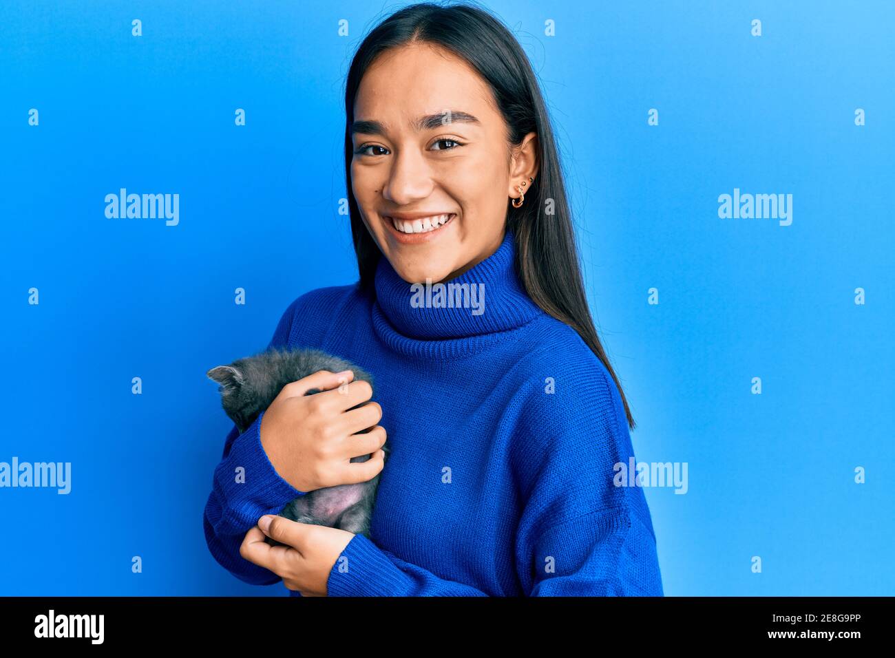 Young hispanic girl smiling happy holding cute cat over isolated blue ...