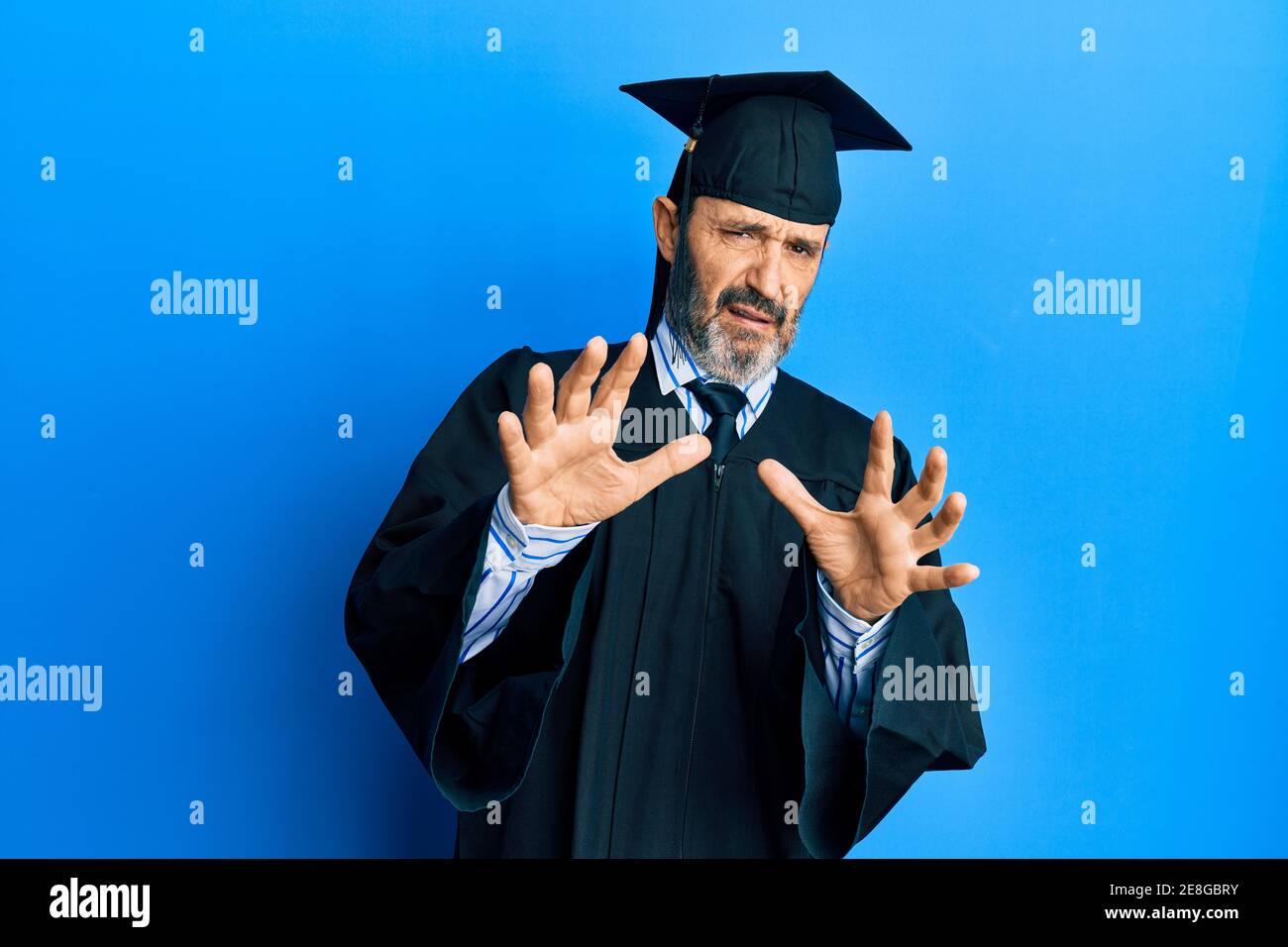 Middle age hispanic man wearing graduation cap and ceremony robe afraid ...
