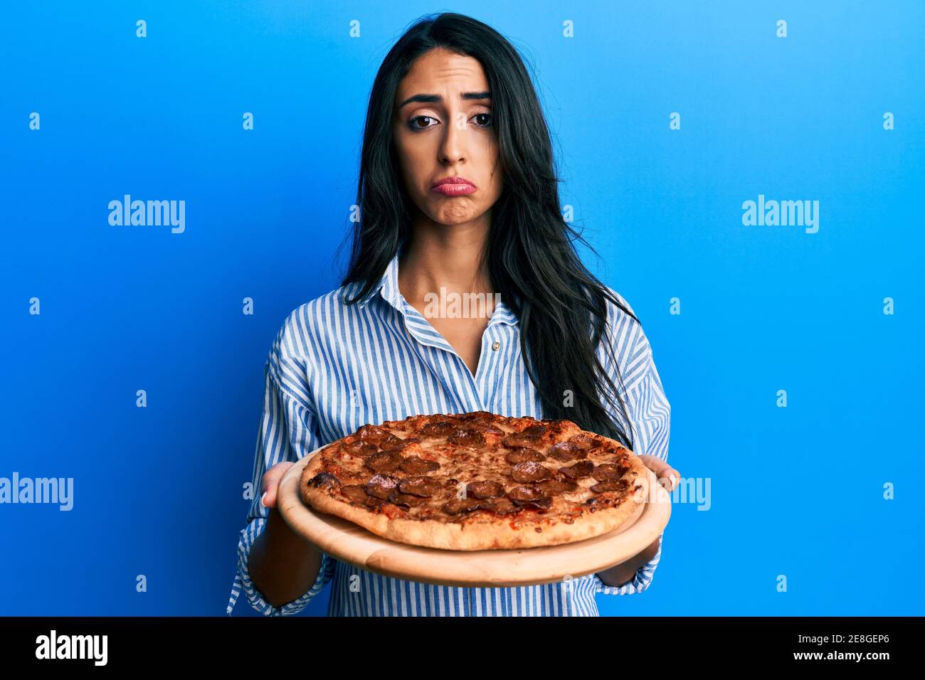 Beautiful hispanic woman holding tasty pepperoni pizza depressed and ...