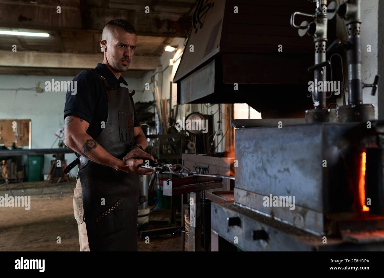 Side view of serious tattooed young male forger in apron heating metal ...