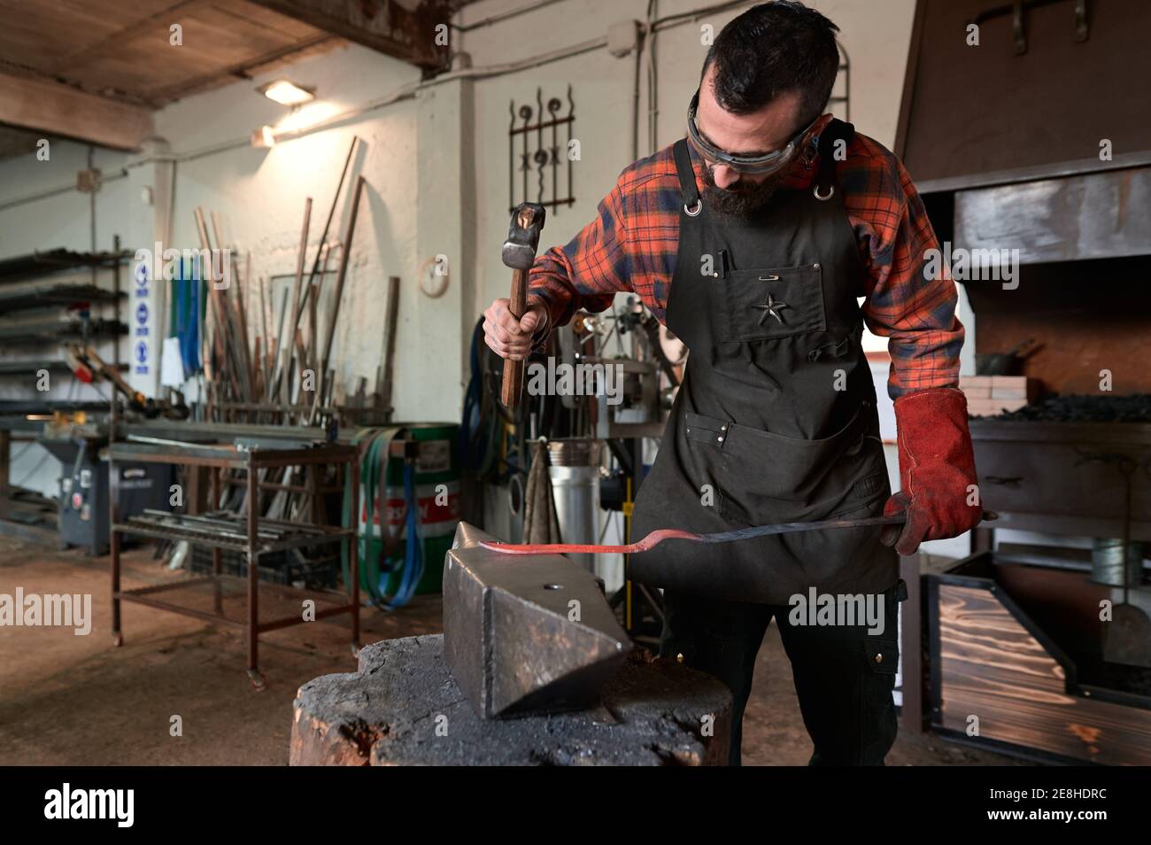 Concentrated young bearded blacksmith in apron and protective goggles ...