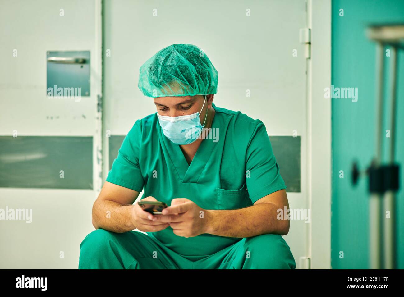 Anonymous young male doctor in green medical uniform and mask messaging ...