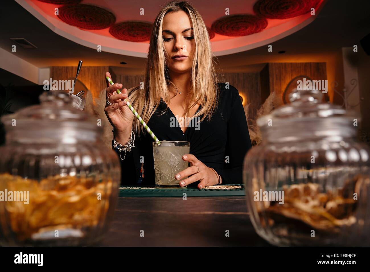 Blonde attractive female bartender stirring iced alcohol cocktail with ...