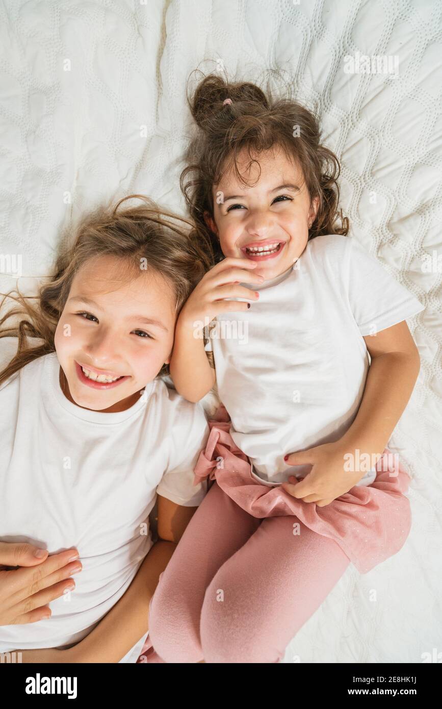 Excited little sisters in casual wear lying on comfy bed together ...