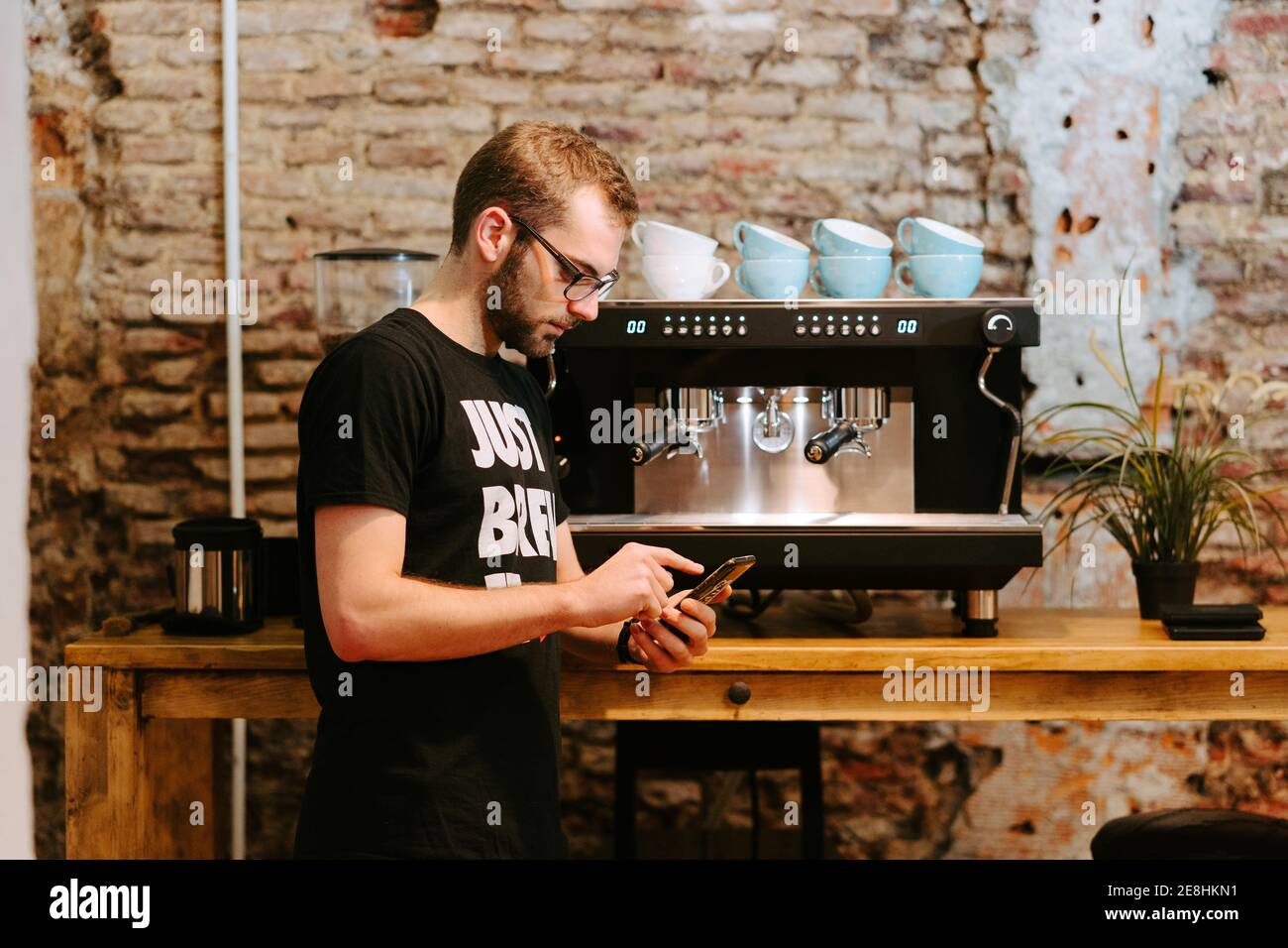 Side view of male barista leaning on wooden counter in coffee shop and ...