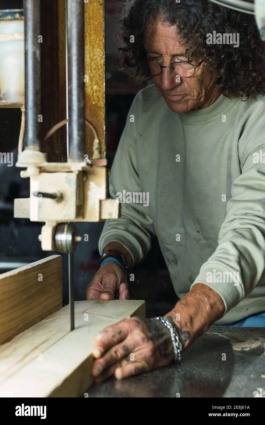 Middle aged male carpenter cutting piece of wood with band saw while ...