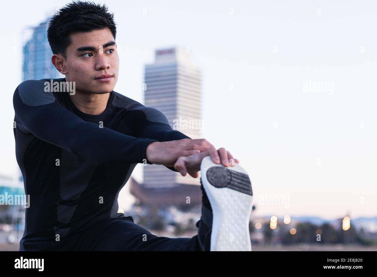 Focused young Asian male in sportswear stretching legs near metal ...