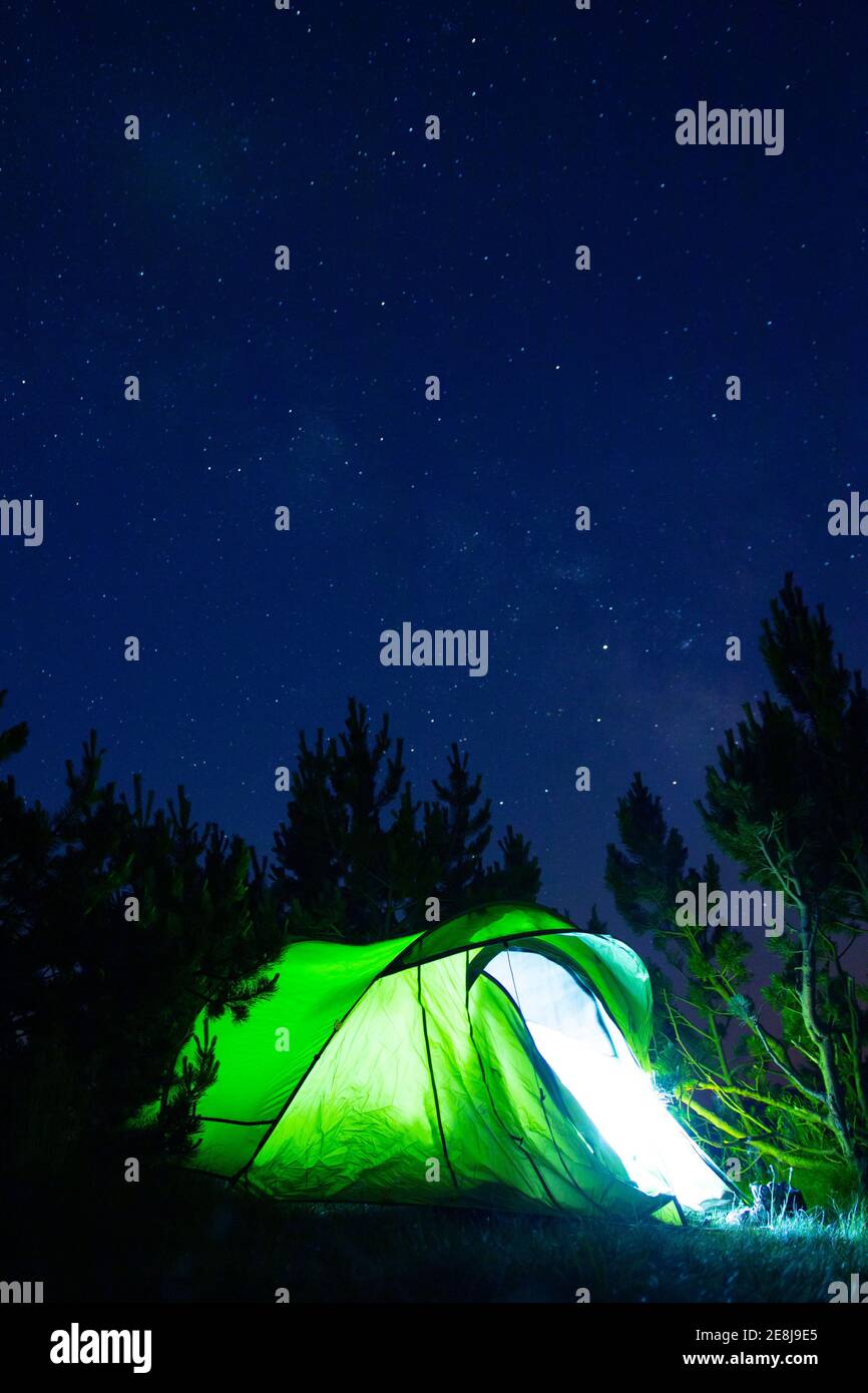 Green tent with shiny light among high tree silhouettes under starry ...