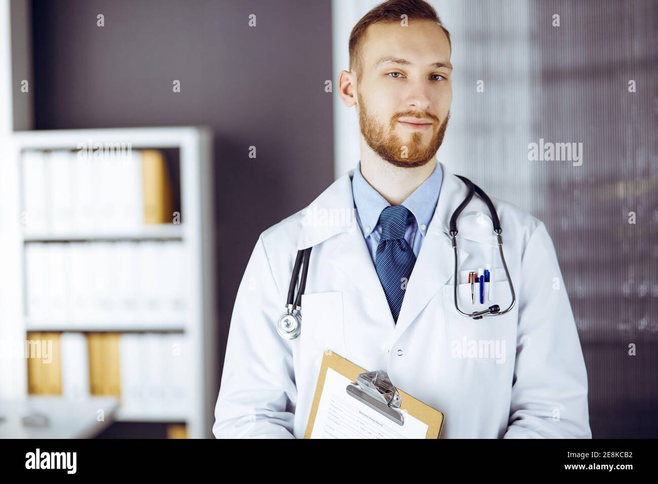 Red-bearded doctor standing straight in clinic near his working place ...