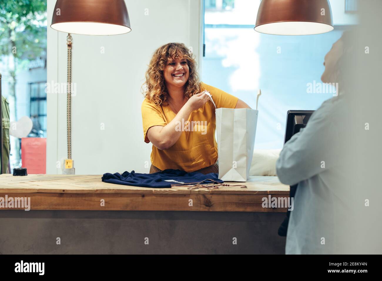 Smiling clothing store owner packing the clothes in a paper bag looking ...