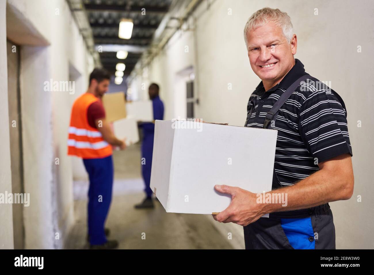 Smiling worker carries cardboard box through an aisle to the mail order ...