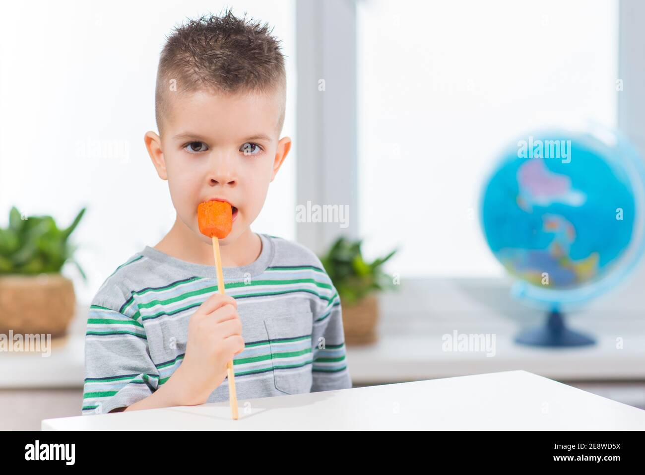 A small child eats a carrot on a stick at home. The boy is eating ...